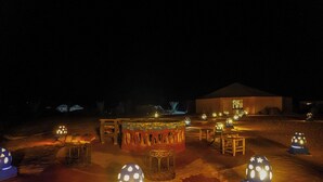 Área externa iluminada à noite, com estruturas esféricas, uma mesa central com toalha de mesa estampada e um edifício ao fundo.