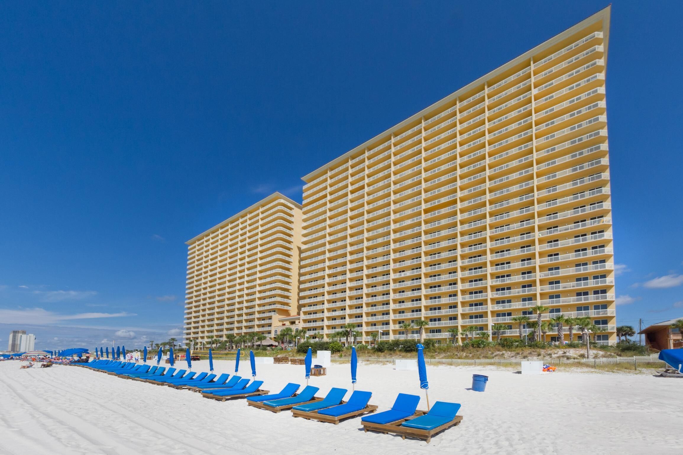 Um hotel na praia com vários edifícios altos, fileiras de espreguiçadeiras azuis e guarda-sóis na areia.