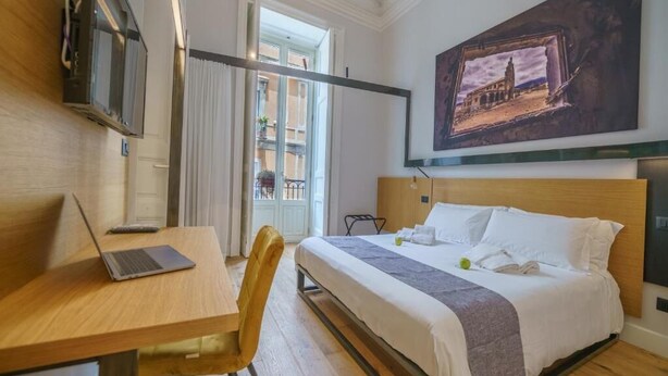 Quarto de hotel com uma cama, uma escrivaninha com um laptop, uma cadeira, uma televisão e um quadro na parede.