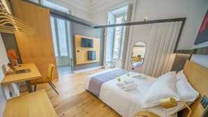 Quarto de hotel moderno com piso de madeira, cama com roupa de cama branca, escrivaninha com cadeira, TV fixada na parede e uma janela grande com cortinas.
