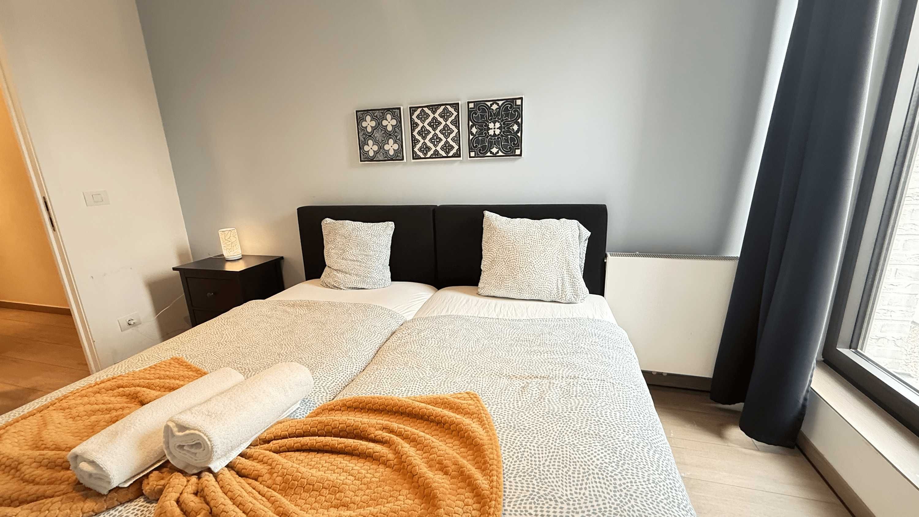 Cama de casal com colcha estampada, dois travesseiros, toalha branca enrolada e manta marrom. Três quadros decoram a parede acima da cama.