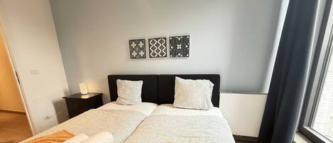 Cama de casal com colcha estampada, dois travesseiros, toalha branca enrolada e manta marrom. Três quadros decoram a parede acima da cama.