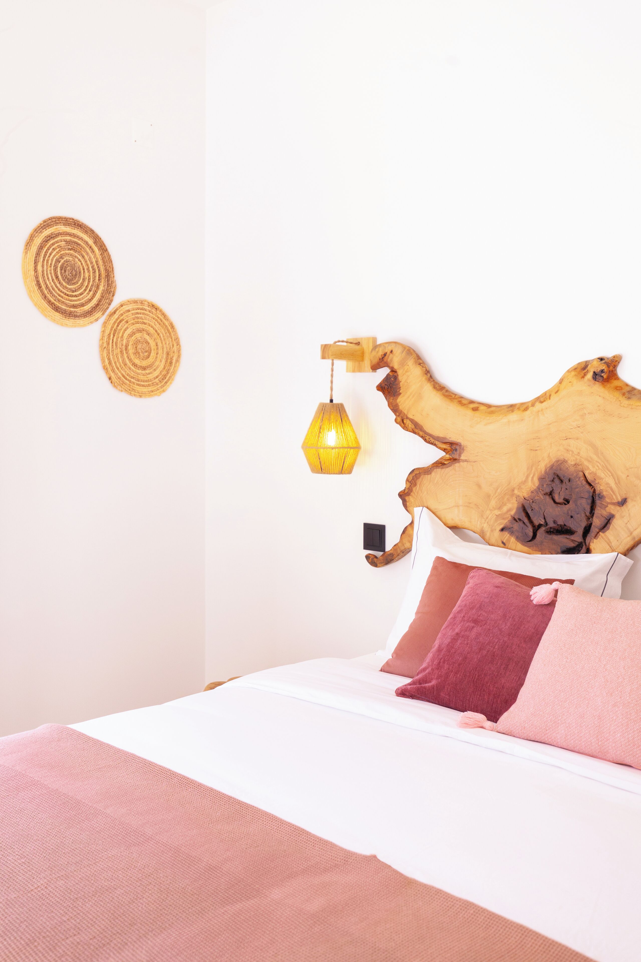 Uma cama com cabeceira de madeira, duas decorações de parede feitas de fibras naturais e um abajur amarelo fixado na parede.