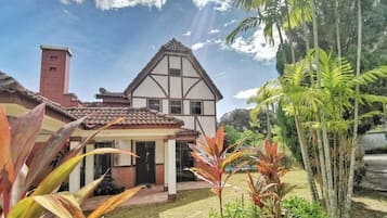 Uma casa com telhado inclinado, chaminé e um jardim bem cuidado com plantas tropicais.