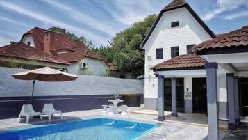 Piscina com espreguiçadeiras, área de estar coberta e um edifício branco com telhado em azulejos.