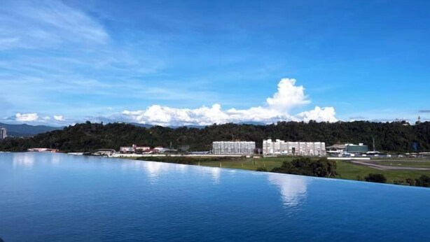 Um vasto corpo de água sob um céu claro, com uma vista distante de edifícios e uma colina arborizada.