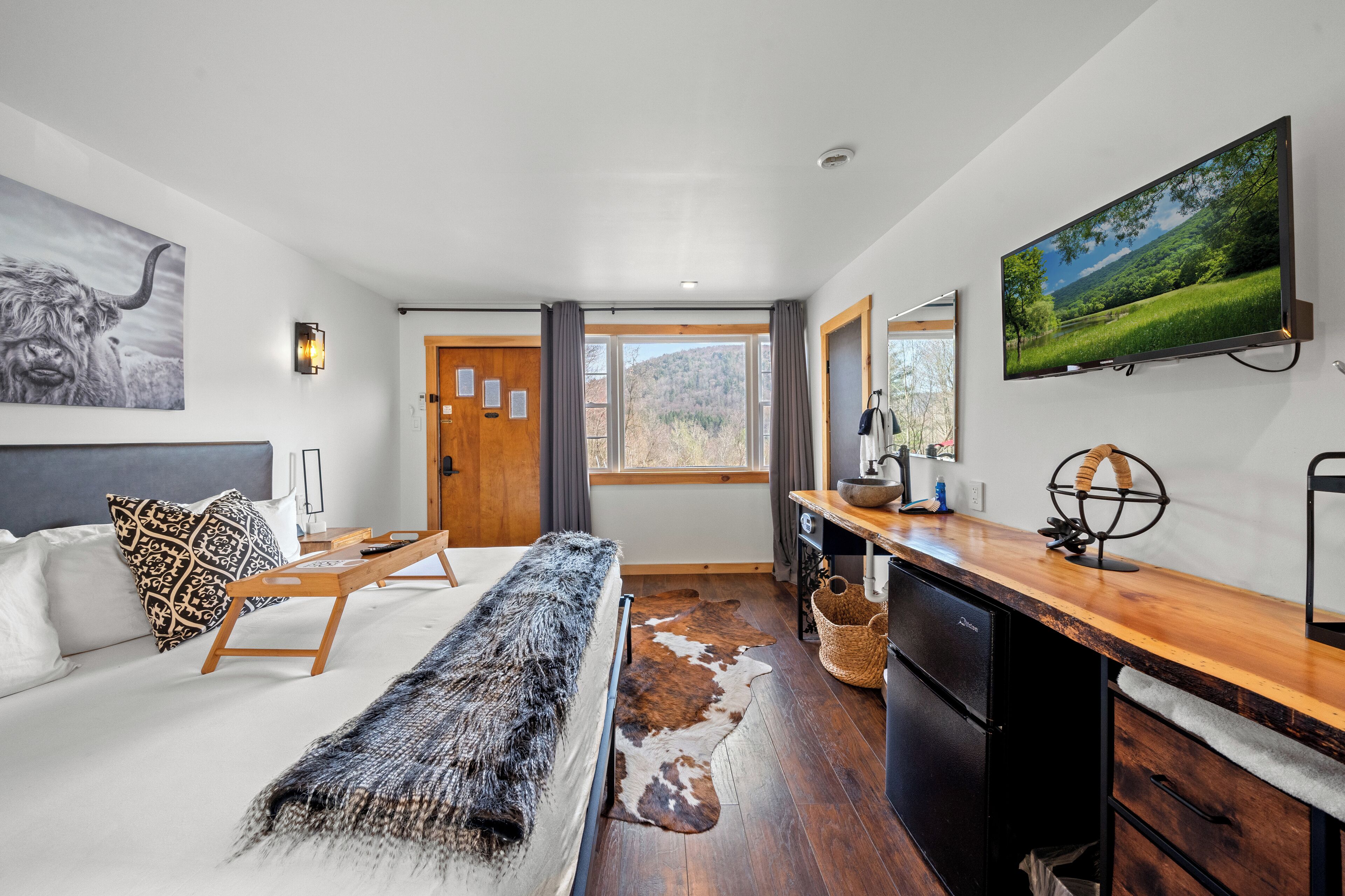 Quarto moderno com cama, escrivaninha de madeira, televisão e uma grande janela com vista para árvores.