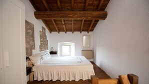 Quarto com teto de madeira, cama com colcha branca, parede de pedra e uma pequena janela.