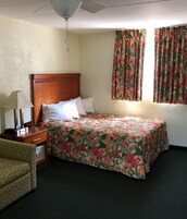 Quarto de hotel com uma cama, mesa de cabeceira, abajur, cadeira e cortinas florais.