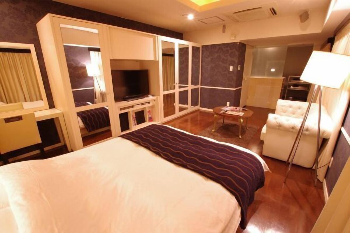 Quarto de hotel com uma cama grande, televisão, área de estar com mesa e cadeiras, e sala separada com sofá e abajur.