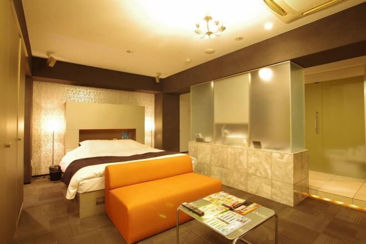 Quarto de hotel com cama, sofá, mesinha com revistas, espelho e área de chuveiro.