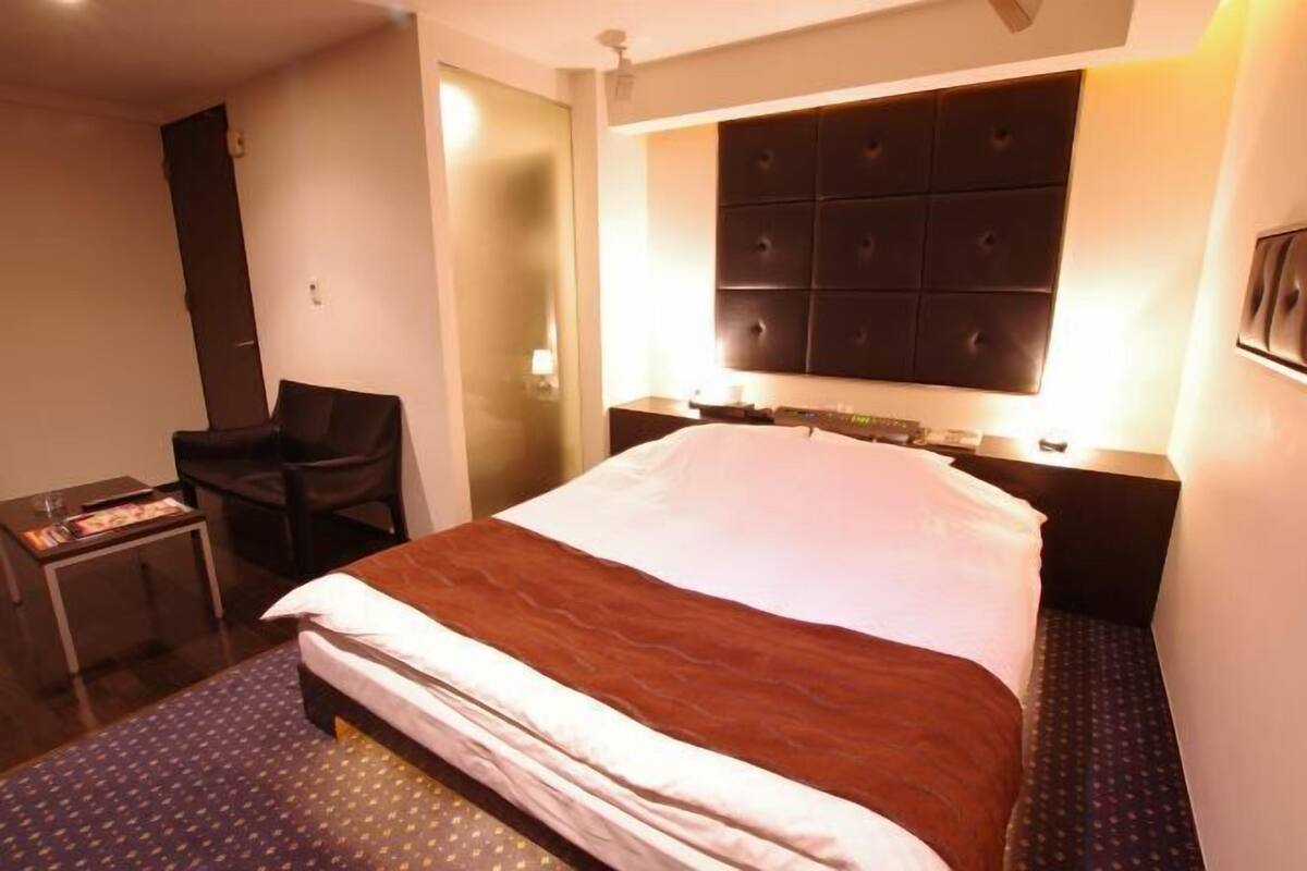 Quarto de hotel com cama grande, mesa de cabeceira, cadeira e cabeceira com iluminação embutida.