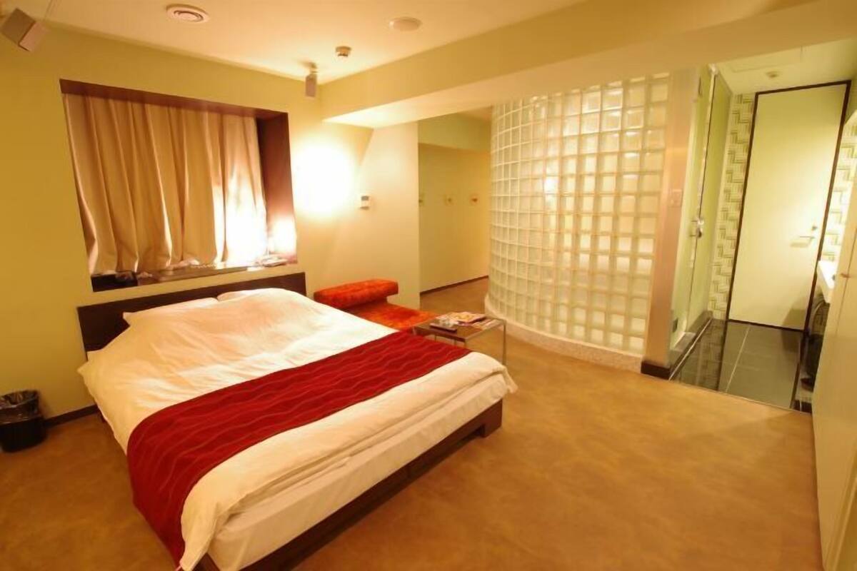 Quarto de hotel com cama, mesinha, cadeira, espelho e divisória de vidro.