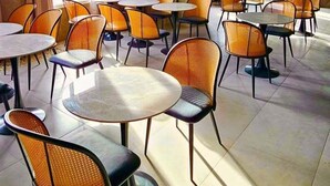 Uma ampla área de refeições interna com várias mesas redondas e cadeiras laranja, grandes janelas com cortinas e um design de teto moderno.