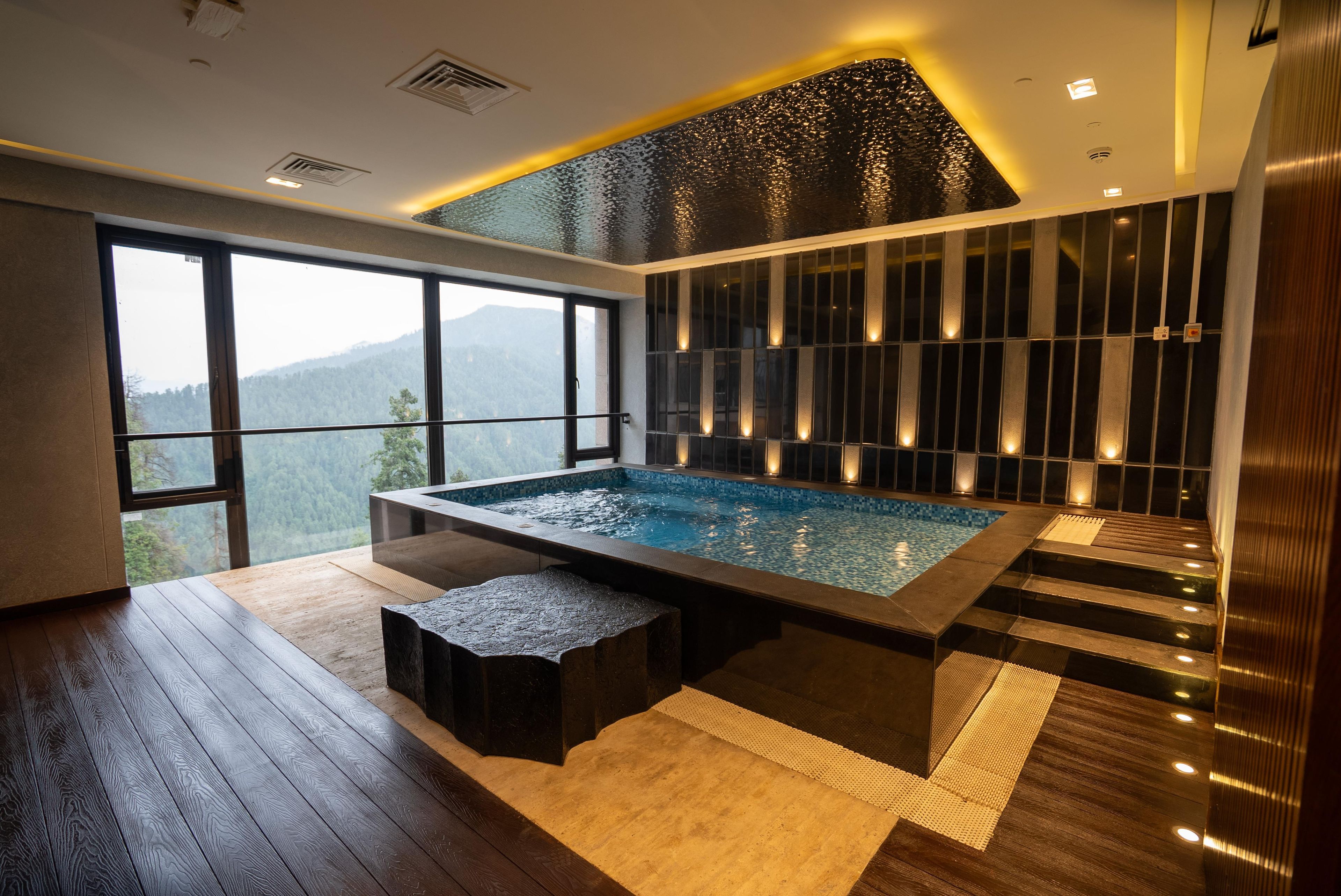 Piscina interna moderna com vista para montanhas, piso de madeira e área de estar.