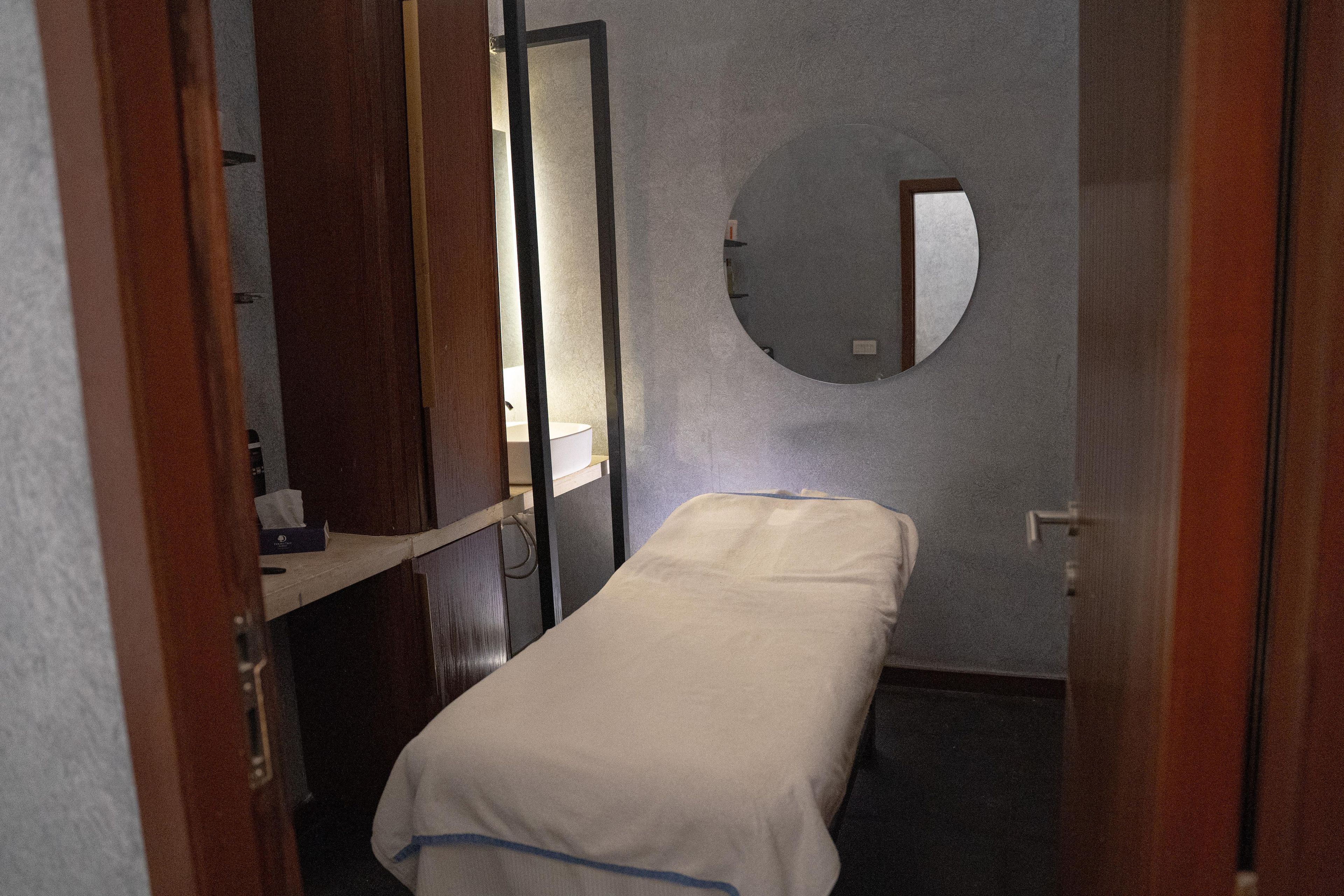 Um quarto pequeno e estreito com uma cama de solteiro, um espelho redondo na parede e uma porta de madeira.