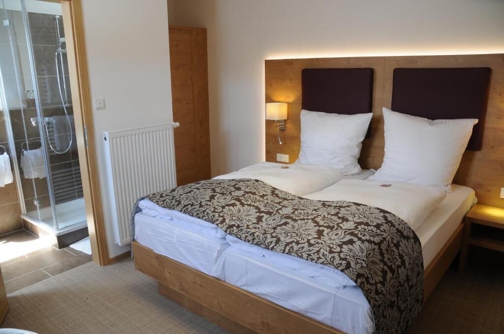 Quarto com cabeceira de madeira, duas luminárias de mesa, cama grande com roupa de cama estampada e box de vidro para o chuveiro.