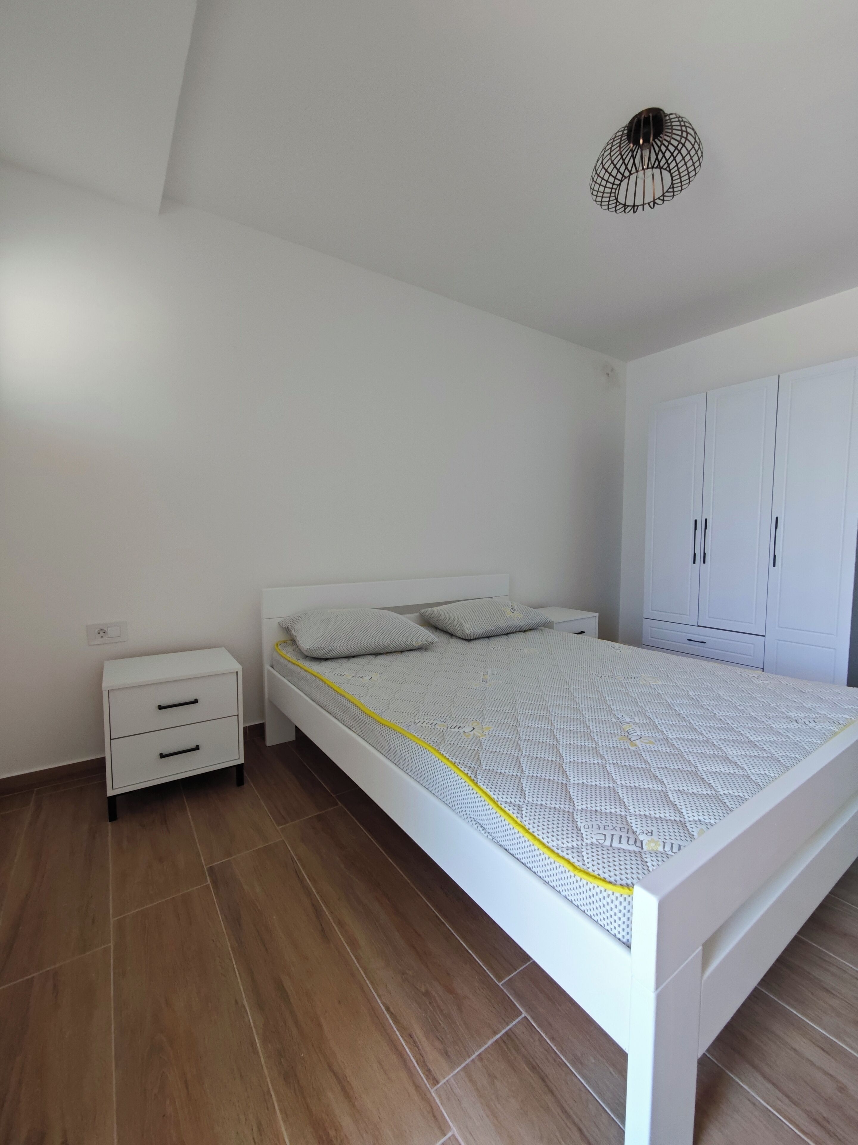 Quarto com cama branca, mesa de cabeceira, guarda-roupa e luminária no teto.