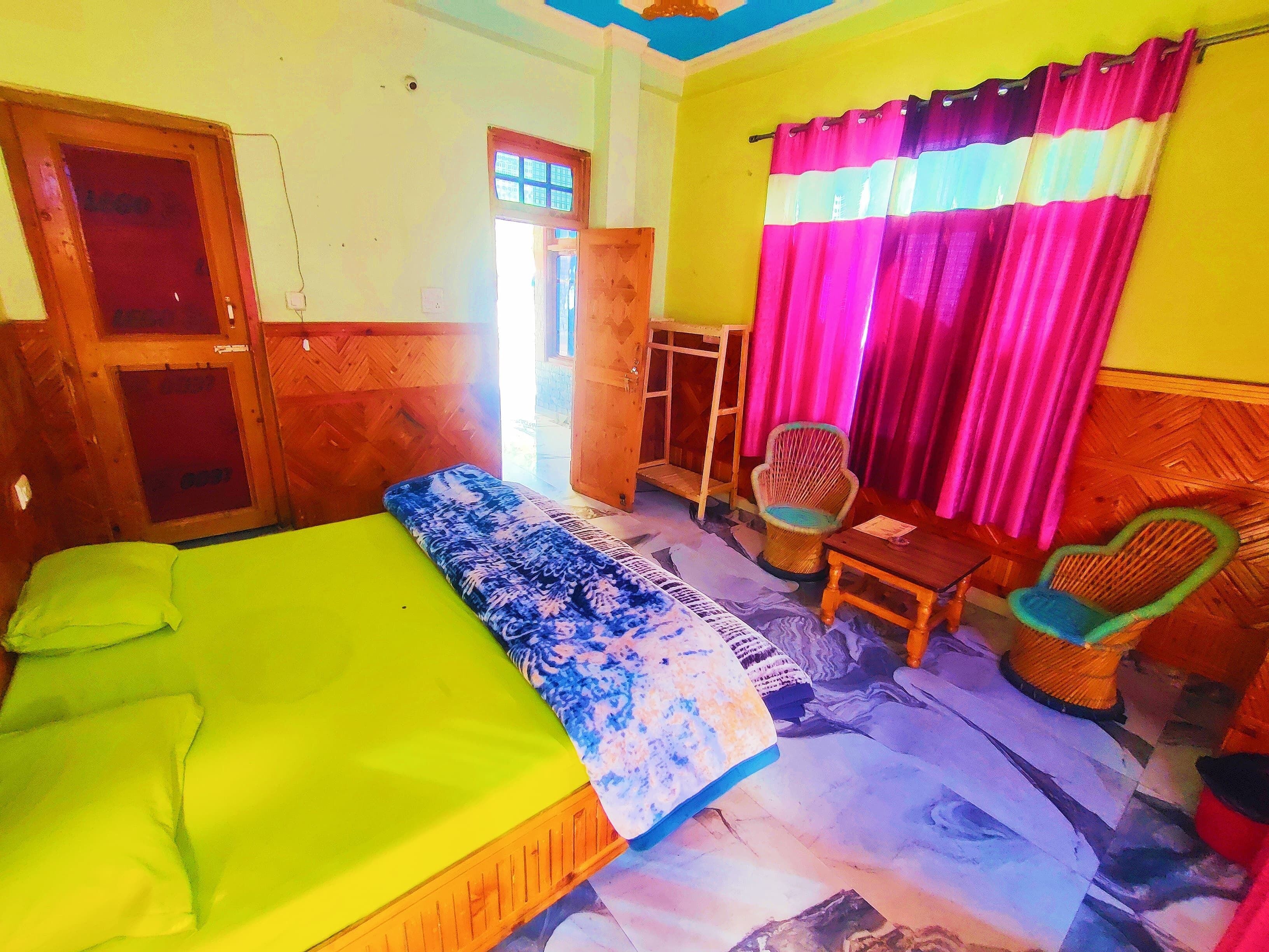 Quarto com cama, duas cadeiras, uma mesinha e janela com cortinas.