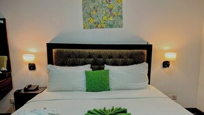 Cama bem arrumada com almofadas brancas e verdes, um cobertor verde e uma toalha dobrada sobre um edredom branco. Há duas luminárias fixadas na parede, uma de cada lado da cama.