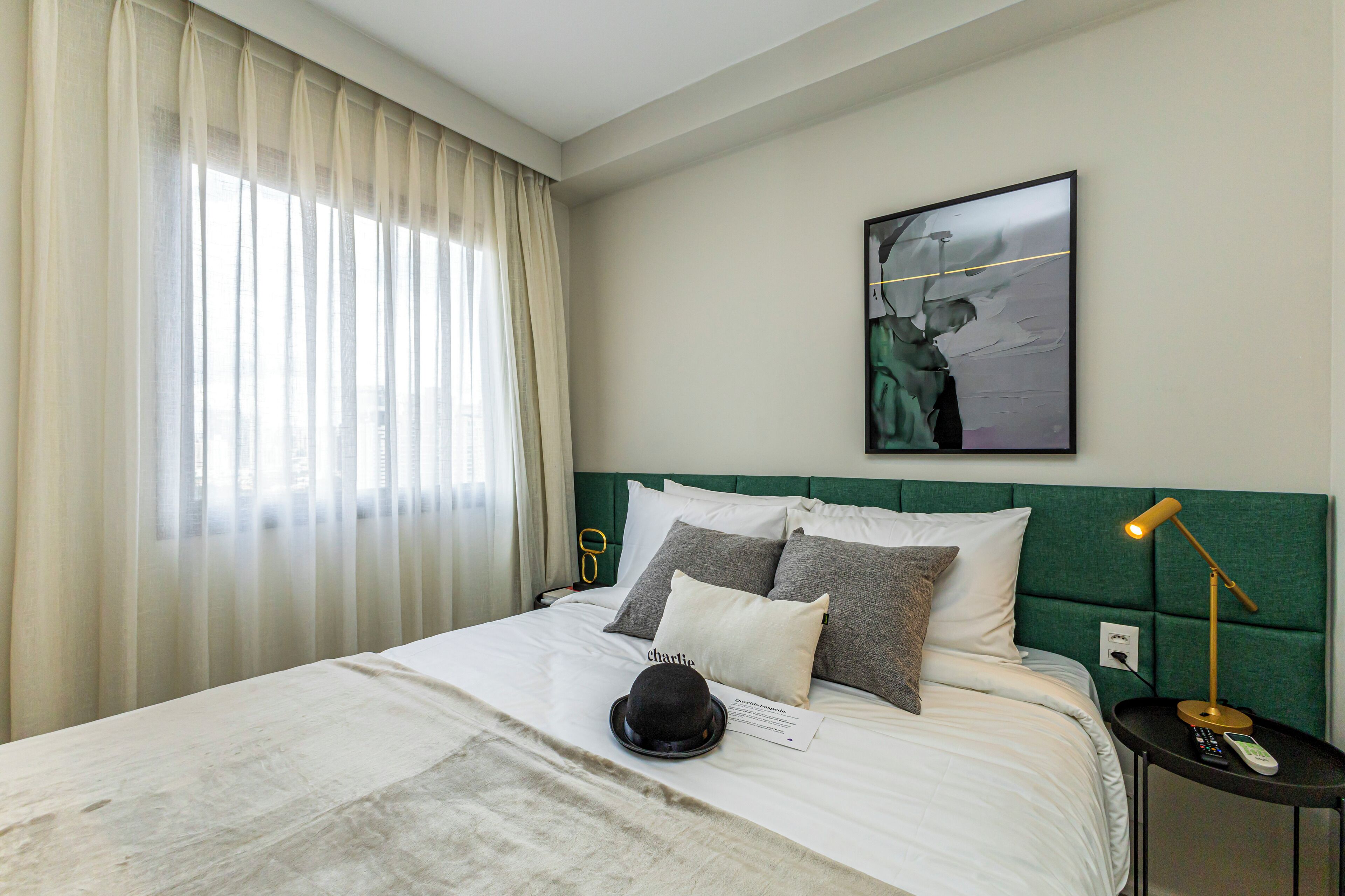 Cama bem arrumada com edredom branco, um chapéu preto e um travesseiro com a palavra "Charlie".  Tem uma cabeceira verde, uma mesinha de cabeceira com abajur e um quadro na parede.