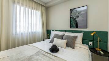 Cama bem arrumada com edredom branco, um chapéu preto e um travesseiro com a palavra "Charlie". Tem uma cabeceira verde, uma mesinha de cabeceira com abajur e um quadro na parede.
