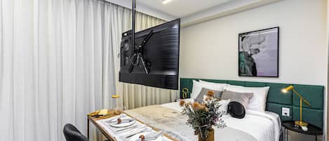 Quarto de hotel moderno com uma mesa de jantar bem organizada, uma grande TV de tela plana na parede, uma cama confortável com cabeceira verde e uma pequena mesa de cabeceira com luminária.