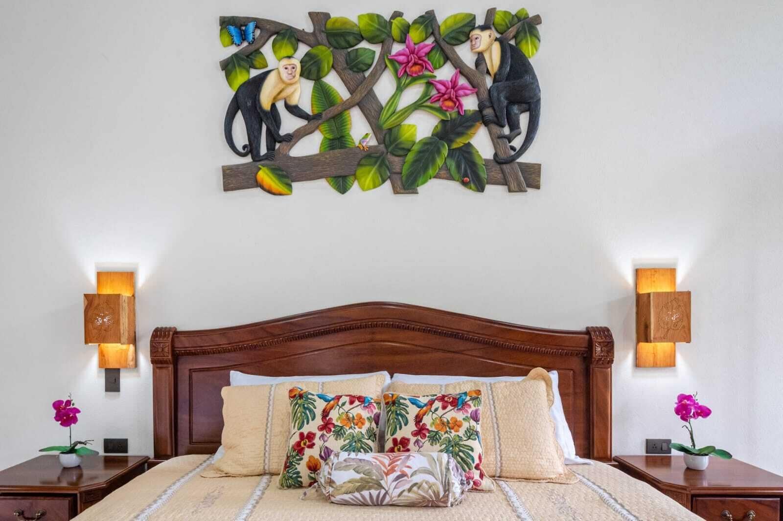 Quarto com cabeceira de madeira, duas luminárias de cabeceira, travesseiro com padrão floral e uma obra de arte na parede retratando macacos em um galho de árvore.