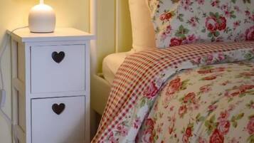 Uma cama bem arrumada com roupa de cama floral, uma luminária de cabeceira e uma mesa de cabeceira branca com puxadores de coração.