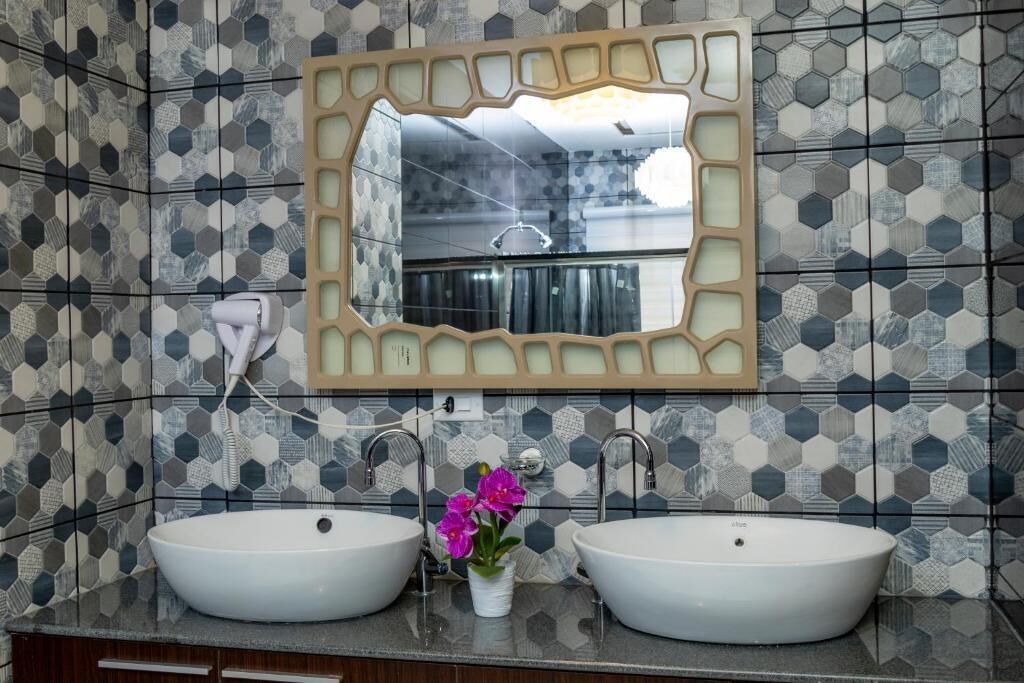 Casa de banho com duas pias brancas, um espelho com moldura geométrica e um painel de azulejos hexagonais estampados.