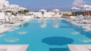 Piscina com cadeiras de lounge e guarda-sóis, cercada por um muro branco e céu azul límpido.