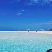 Uma praia com águas azul-claras e areia branca. Há guarda-sóis e pessoas na praia.