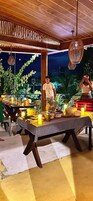 Uma área de refeições ao ar livre com uma mesa posta para dois, rodeada por vigas de madeira e luminárias pendentes.