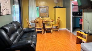 Uma sala de estar com sofá de couro preto, mesa de jantar de madeira com cadeiras, piso de madeira, ventilador de teto e uma área de cozinha com televisão.