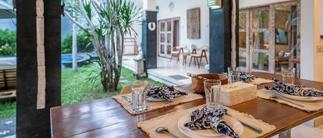 Uma mesa de jantar posta com pratos, copos e talheres, com vista para um jardim e área de estar ao ar livre.