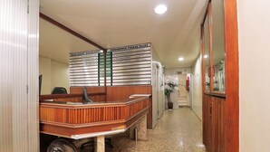 Corredor de hotel com balcão de recepção de madeira, um ventilador e um mural com números e nomes de quartos.