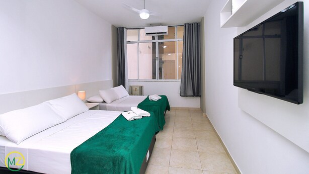 Quarto de hotel com duas camas, ventoinha no teto, ar condicionado, televisão fixada na parede e janela com cortinas.