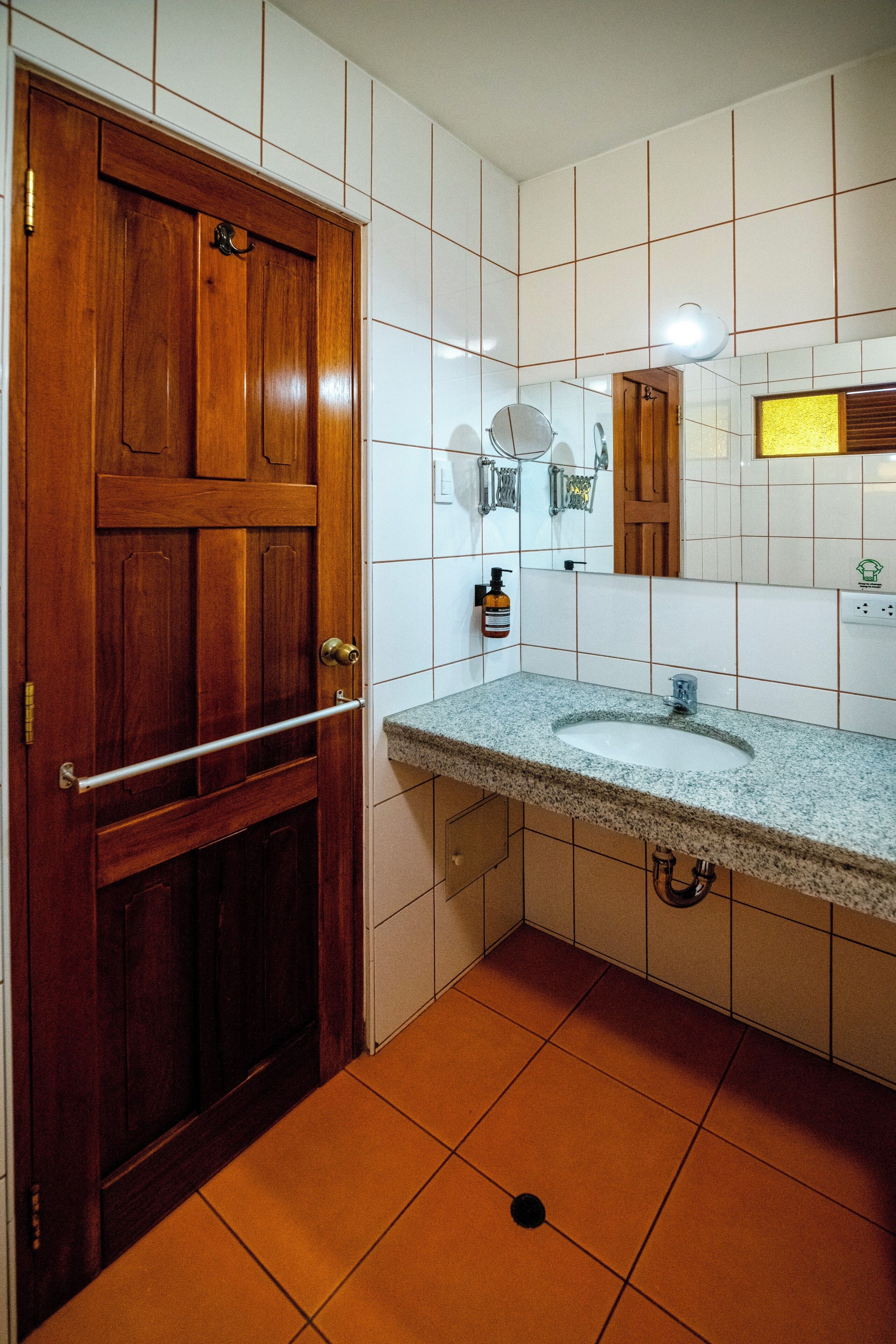 Uma casa de banho com porta de madeira, paredes com azulejos brancos, lavatório com bancada de granito e espelho em cima.