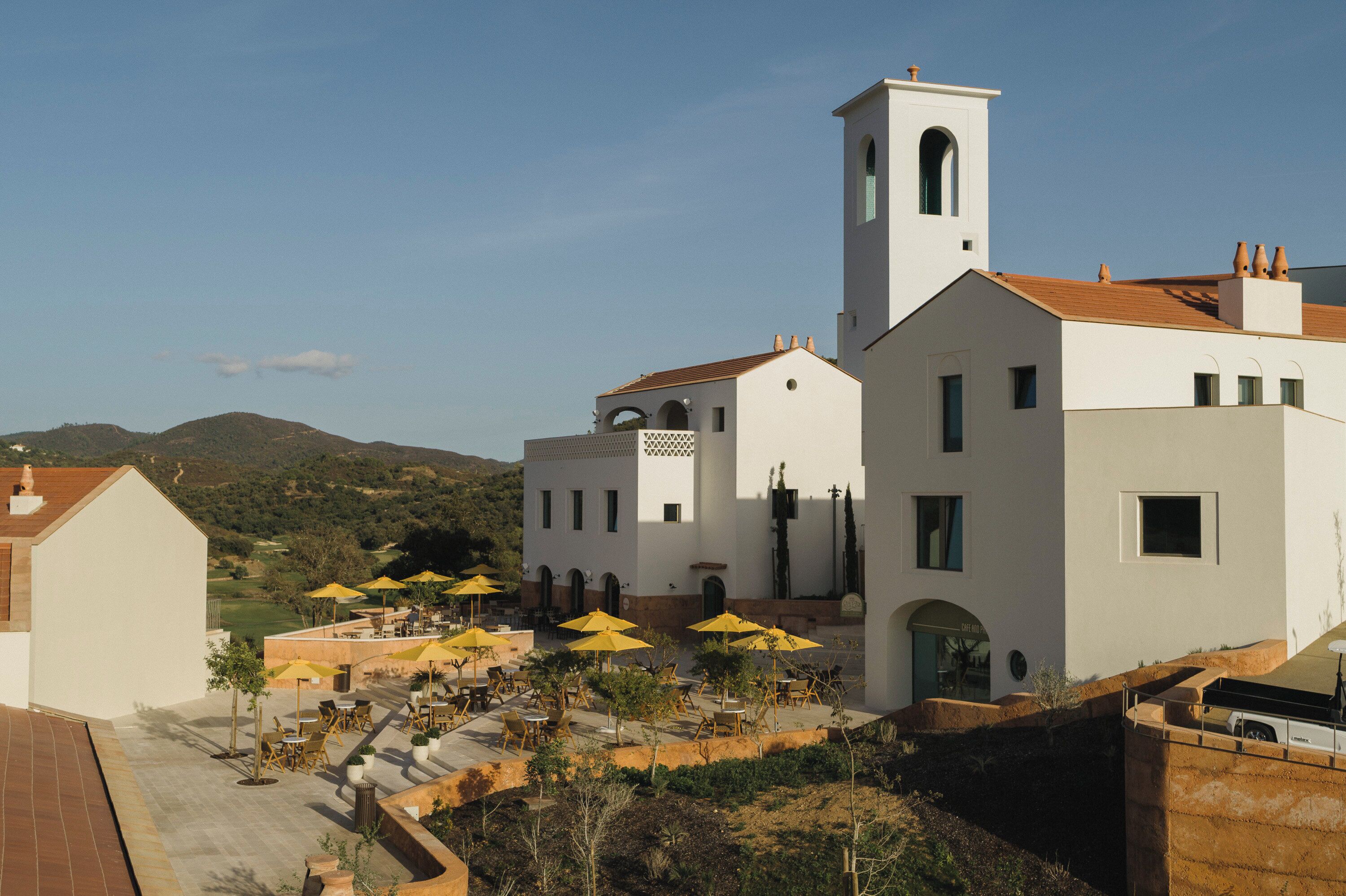 Um edifício com uma torre, um pátio com mesas e guarda-sóis amarelos, e montanhas ao fundo.