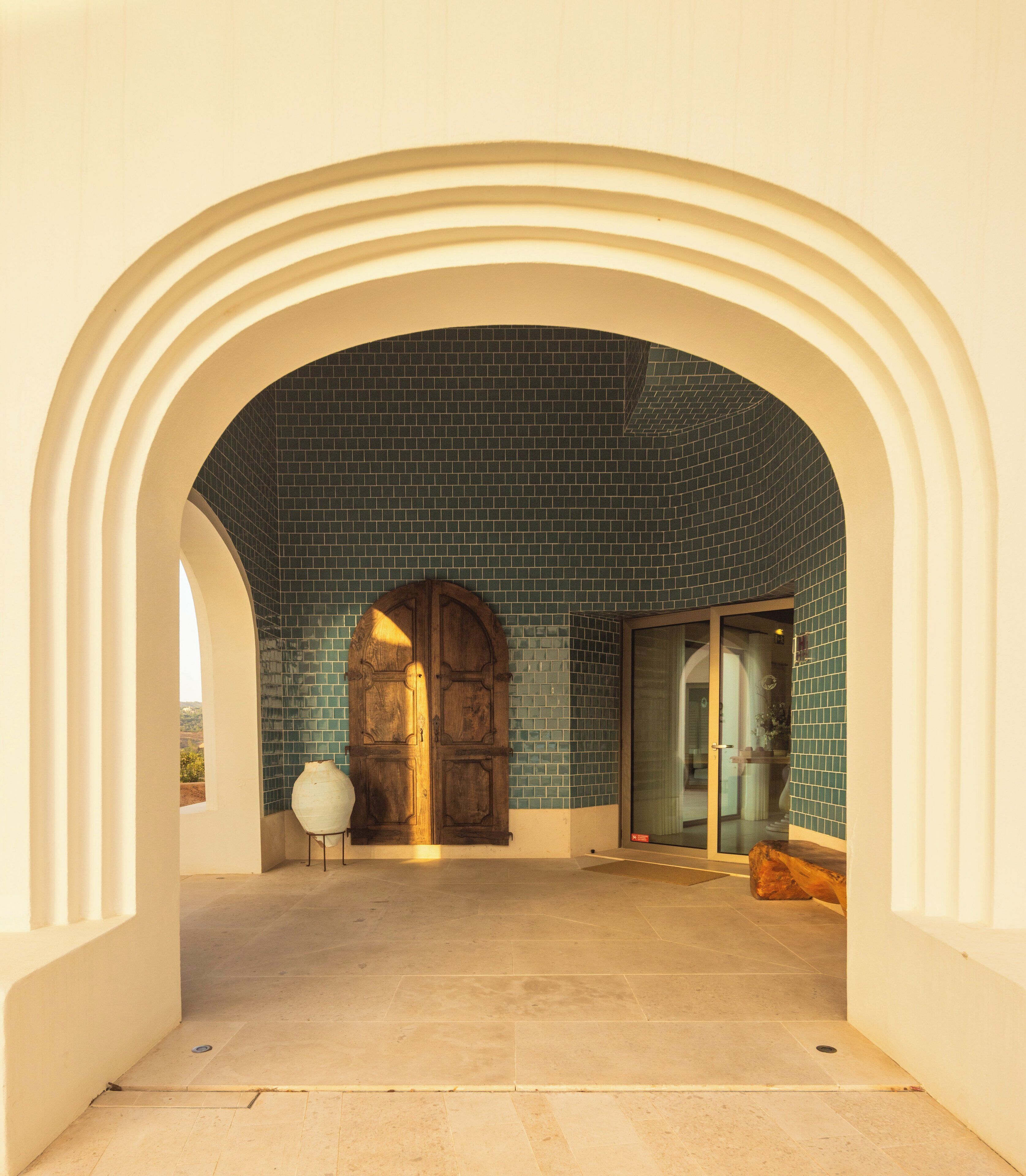 Uma entrada arqueada com um muro revestido de azulejos, uma porta de madeira e uma porta de vidro que dá para o interior.