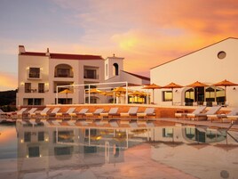 Área à beira da piscina com espreguiçadeiras e guarda-sóis, refletindo o edifício e o céu.