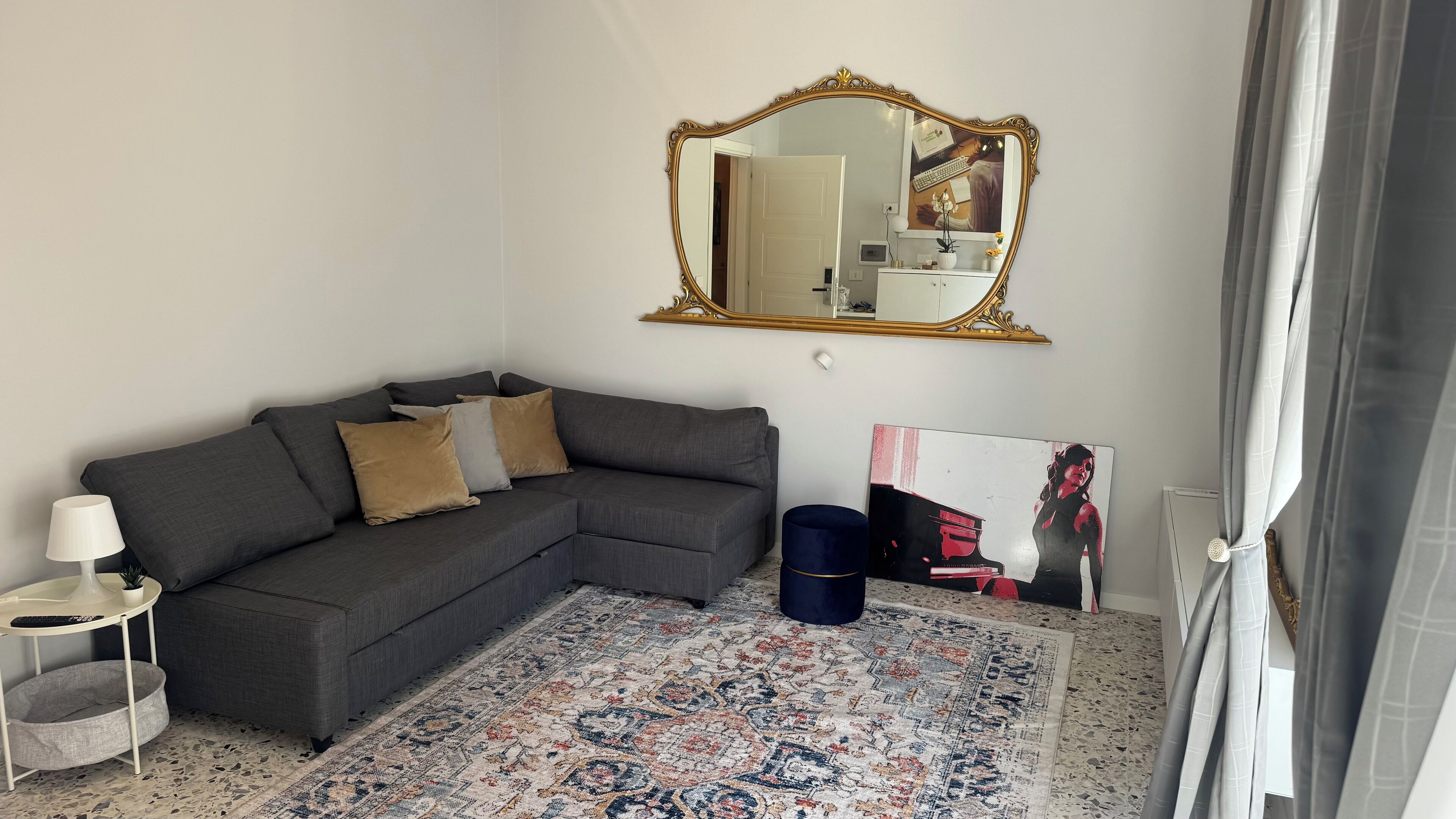 Uma sala de estar com um sofá modular cinza, um tapete estampado, uma mesinha de apoio com abajur e um espelho grande na parede.