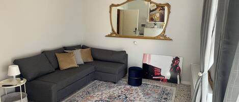 Uma sala de estar com um sofá modular cinza, um tapete estampado, uma mesinha de apoio com abajur e um espelho grande na parede.
