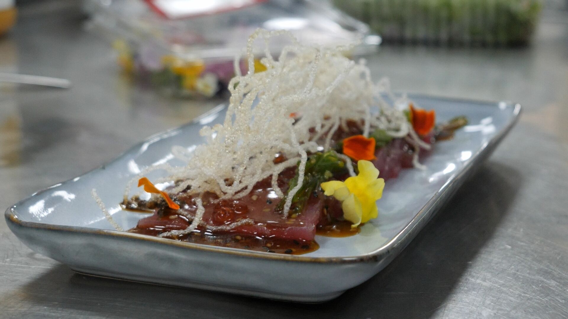 Um prato quadrado com uma porção de sashimi, decorado com cobertura branca desfiada, legumes e molho escuro.