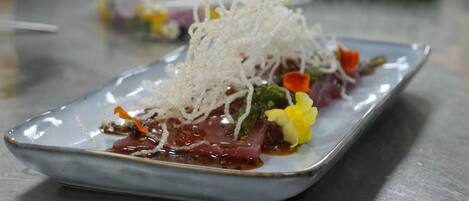 Um prato quadrado com uma porção de sashimi, decorado com cobertura branca desfiada, legumes e molho escuro.