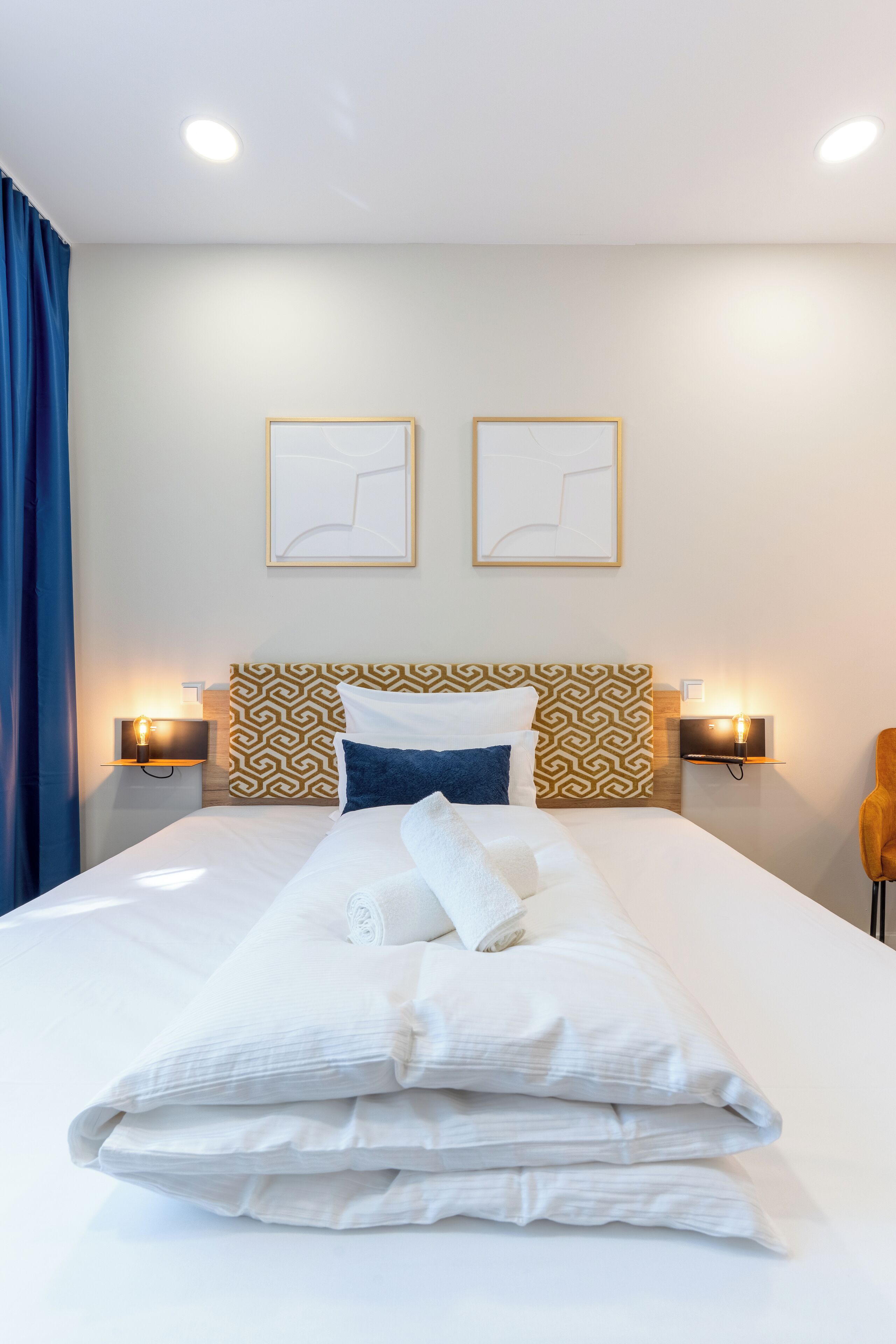 Uma cama bem arrumada com lençóis brancos e uma toalha dobrada, duas obras de arte emolduradas na parede e uma mesa de cabeceira com uma luminária.