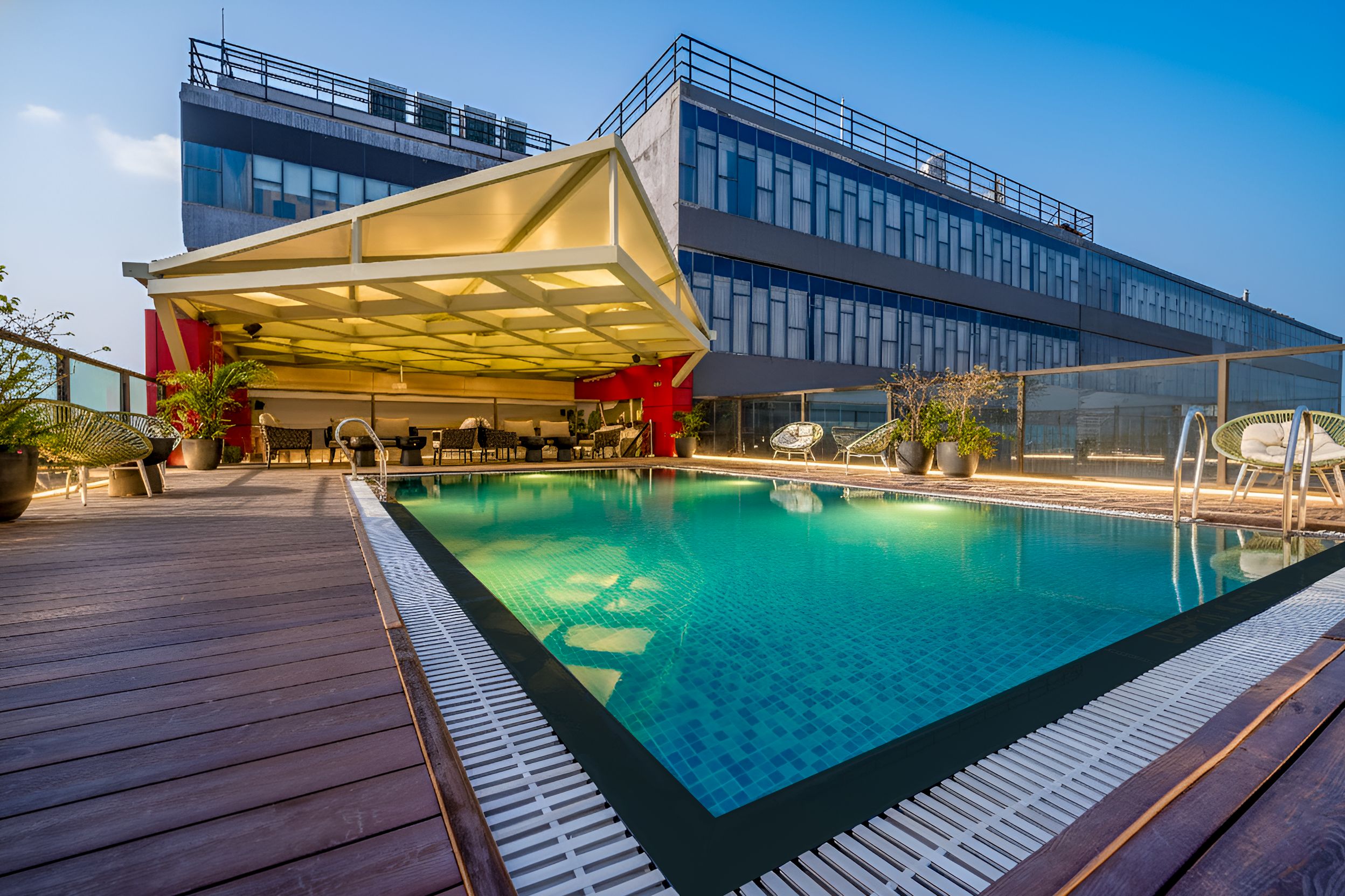 Piscina com deck de madeira, área de estar coberta e um edifício moderno ao fundo.