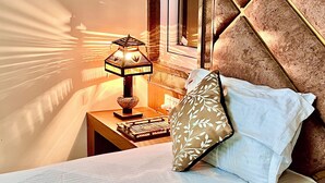 Cama bem arrumada com lençóis brancos e almofada decorativa com padrão de folha. Criado-mudo de madeira acomoda uma luminária de mesa moderna com design geométrico.