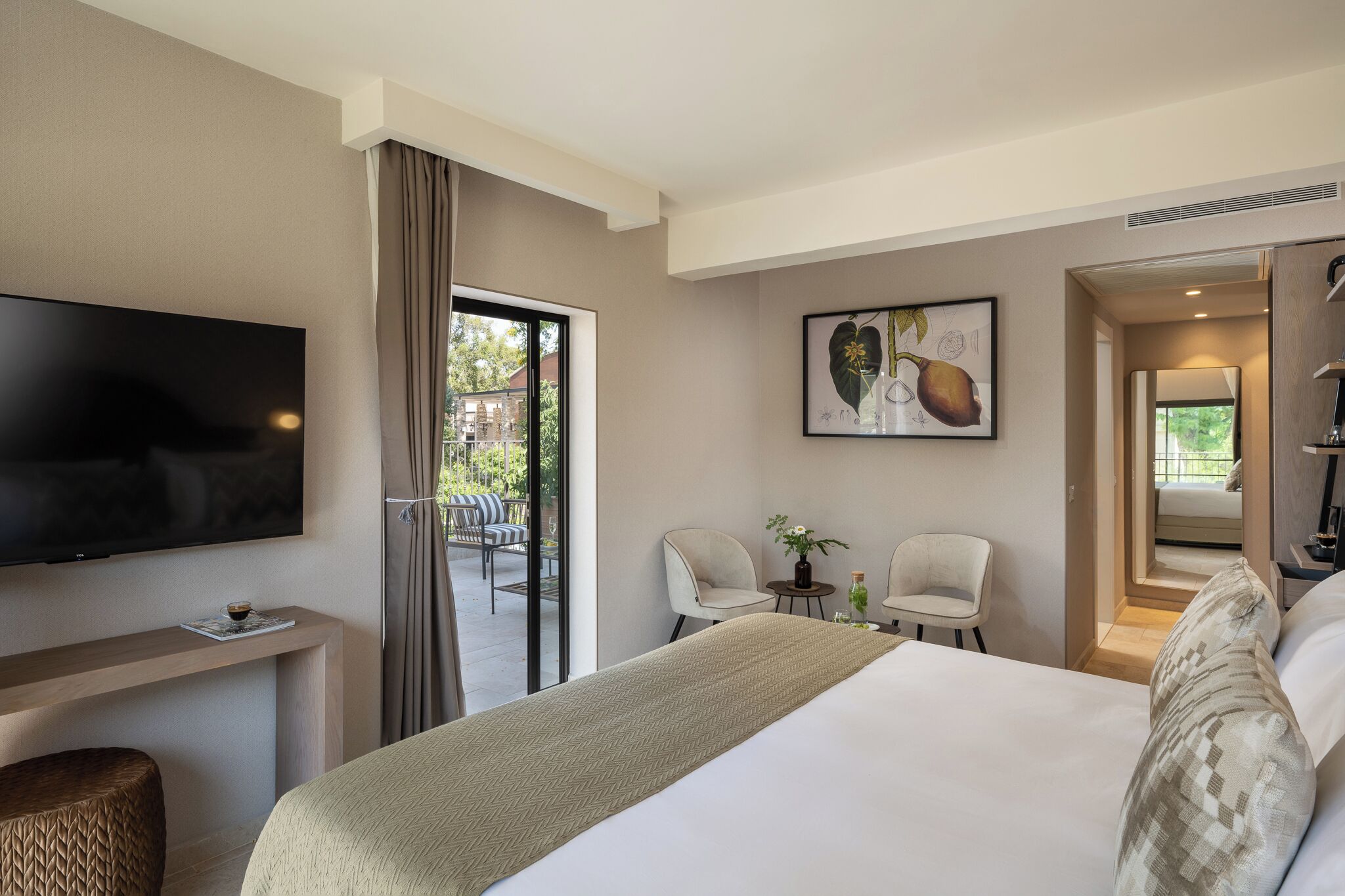Quarto de hotel moderno com uma cama grande, televisão de tela plana, uma mesinha com um vaso e uma cadeira.
