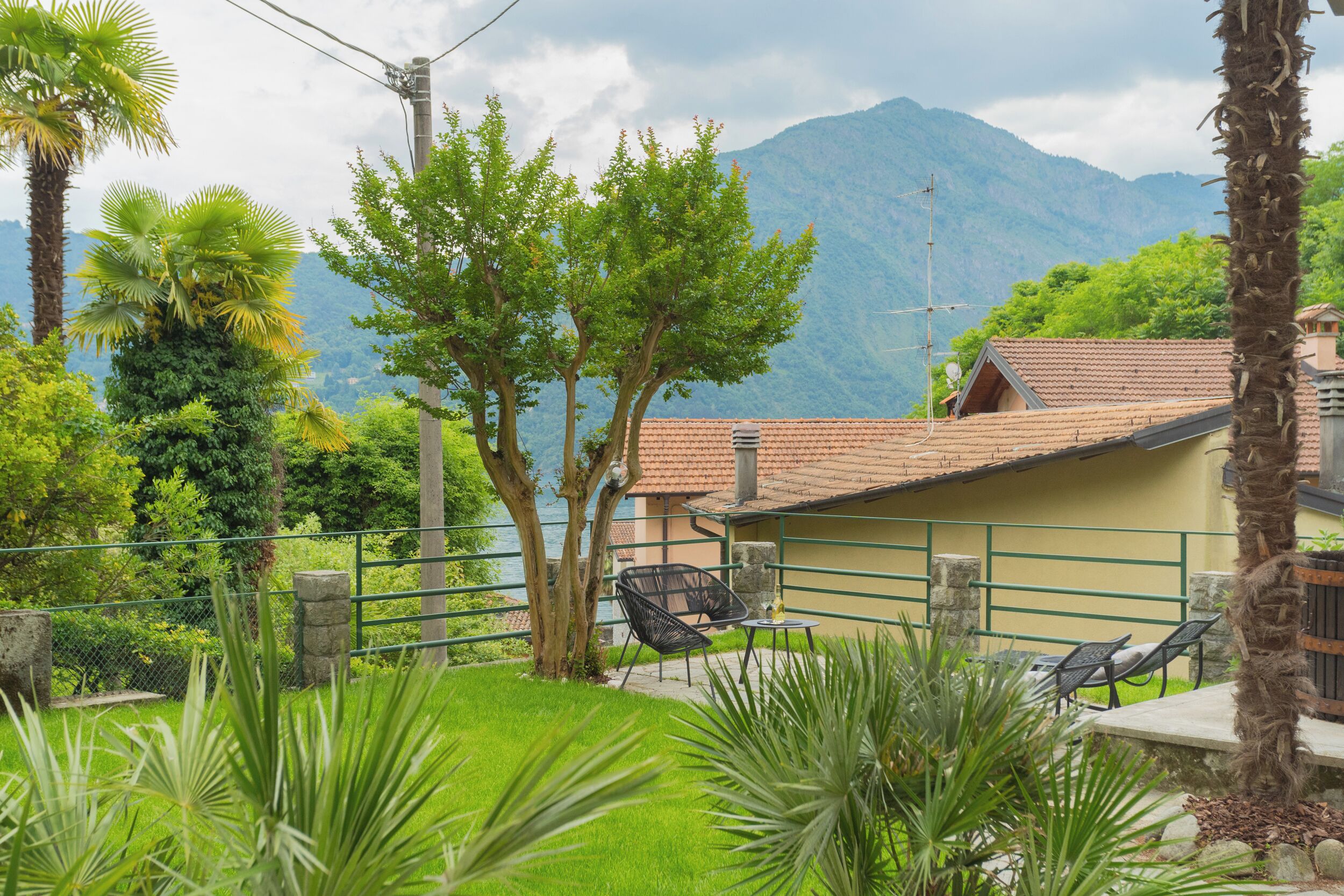 Uma casa com telhado de telhas, jardim cercado e montanha ao fundo.
